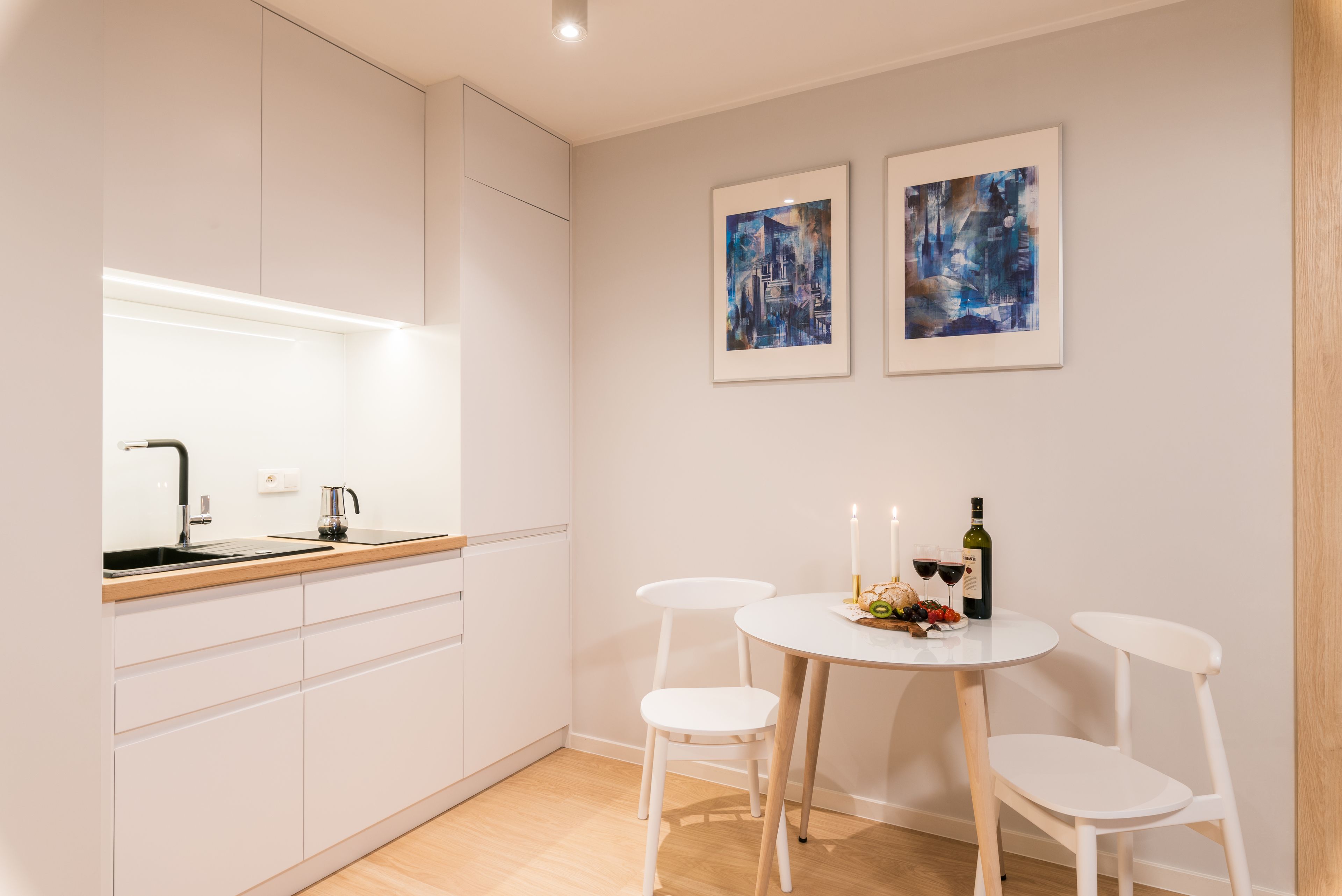 Uma cozinha moderna com armários brancos, bancada de madeira e um espaço de refeições com mesa redonda e cadeiras brancas. Há duas pinturas abstratas emolduradas na parede.