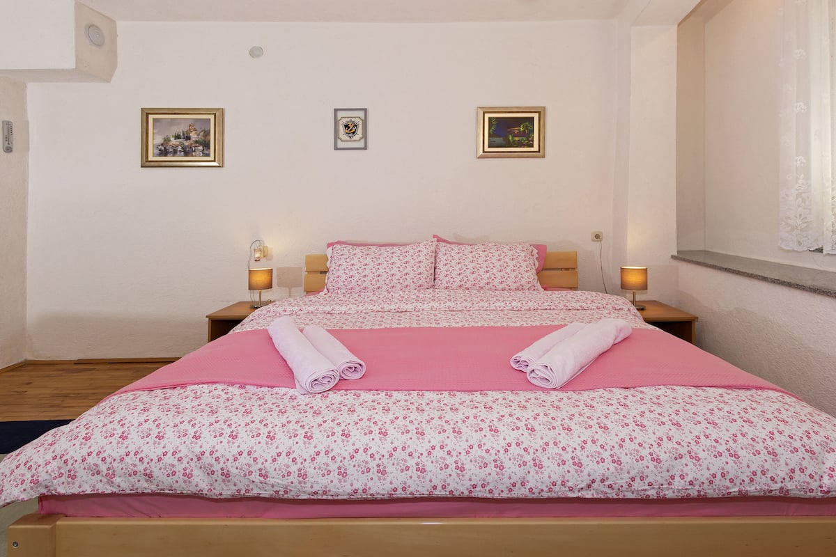 Uma cama de casal com roupa de cama floral em rosa e branco, duas luminárias de cabeceira e três quadros na parede.