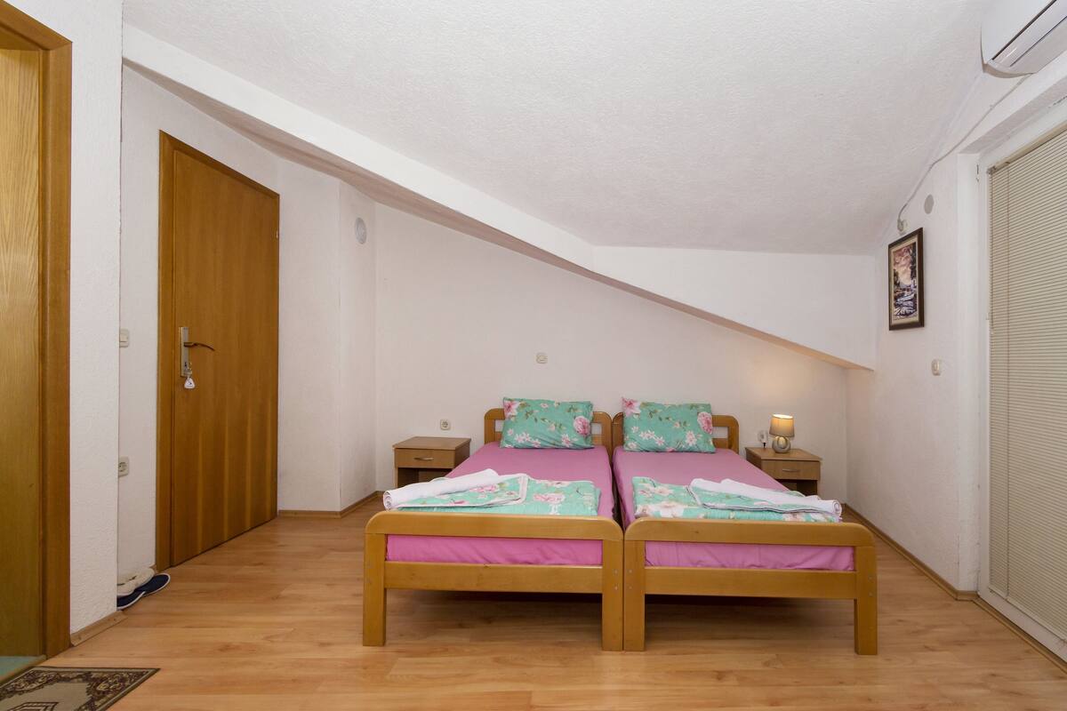 Quarto com duas camas de madeira, uma mesinha de cabeceira, ar-condicionado fixado na parede e uma porta.