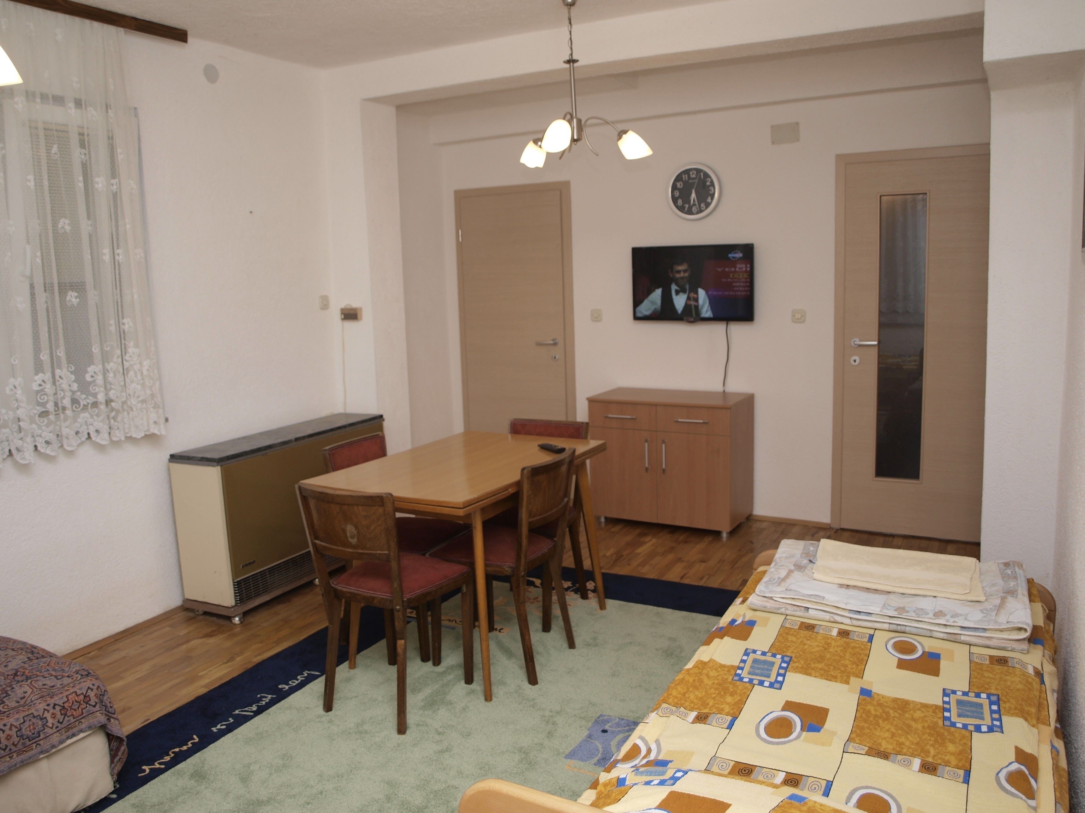 Um quarto pequeno e acolhedor, com uma mesa de jantar, cadeiras, televisão na parede e uma cama com colcha estampada.