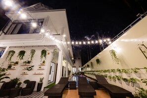 Um hall de hotel moderno com piso xadrez, poltronas de vime, piscina e plantas em vasos nas prateleiras e paredes.