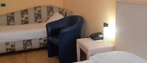 Quarto de hotel com uma cama, uma cadeira, uma mesinha com telefone e abajur, e um sofá estampado com flores.