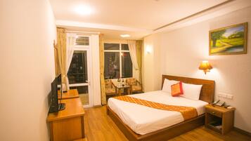 Quarto de hotel com cama de madeira, uma escrivaninha com computador, uma mesa pequena com cadeiras e uma janela com cortinas.
