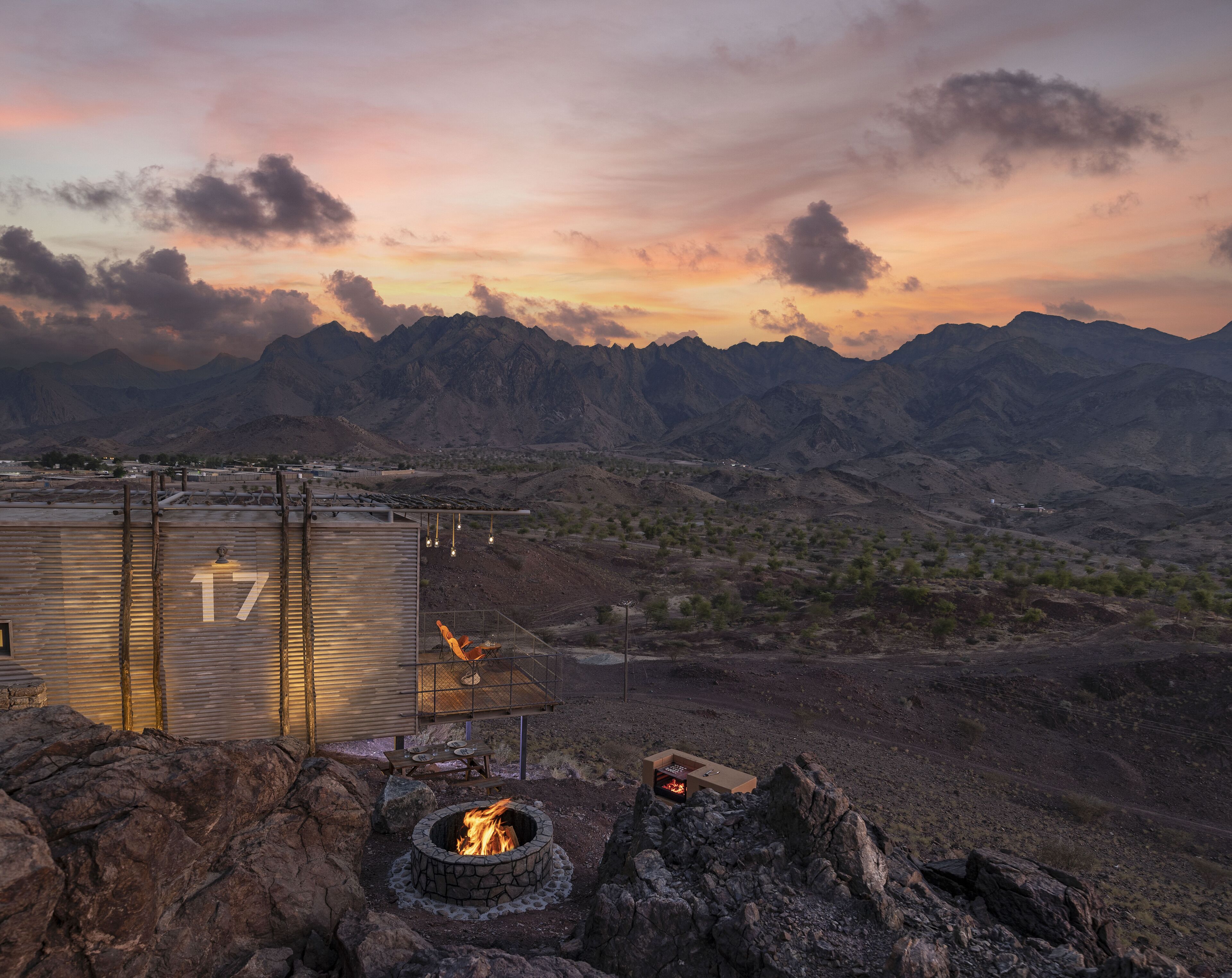 Uma cabana rústica com um poço de fogo de pedra, um deck com luzes penduradas e vista para montanhas ao fundo.