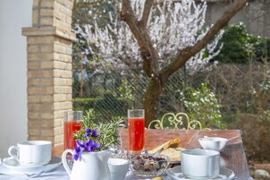 Disposição para o café da manhã, com uma mesa, xícaras, jarra e um copo de bebida vermelha, em frente a uma parede de tijolos e uma árvore com flores brancas.