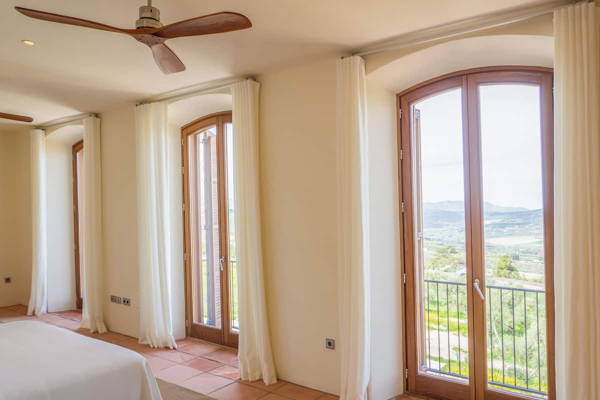 Quarto com cama, ventilador de teto e amplas janelas com cortinas.