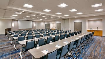 Uma sala de conferências com fileiras de mesas e cadeiras, uma tela de projetor e uma pequena copa.