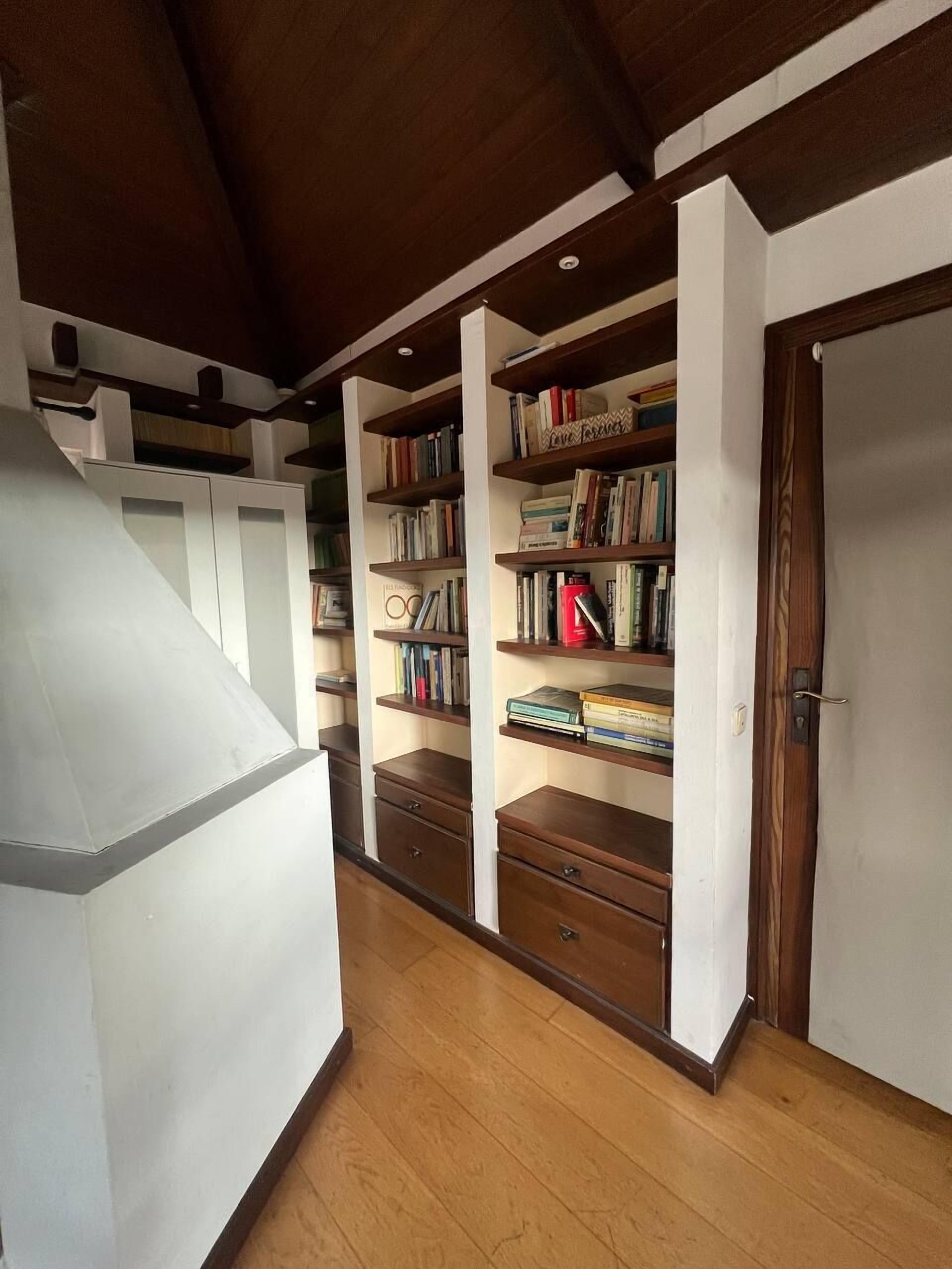 Quarto com piso de madeira, paredes brancas e uma estante repleta de livros. Há uma porta à direita e um armário branco à esquerda.