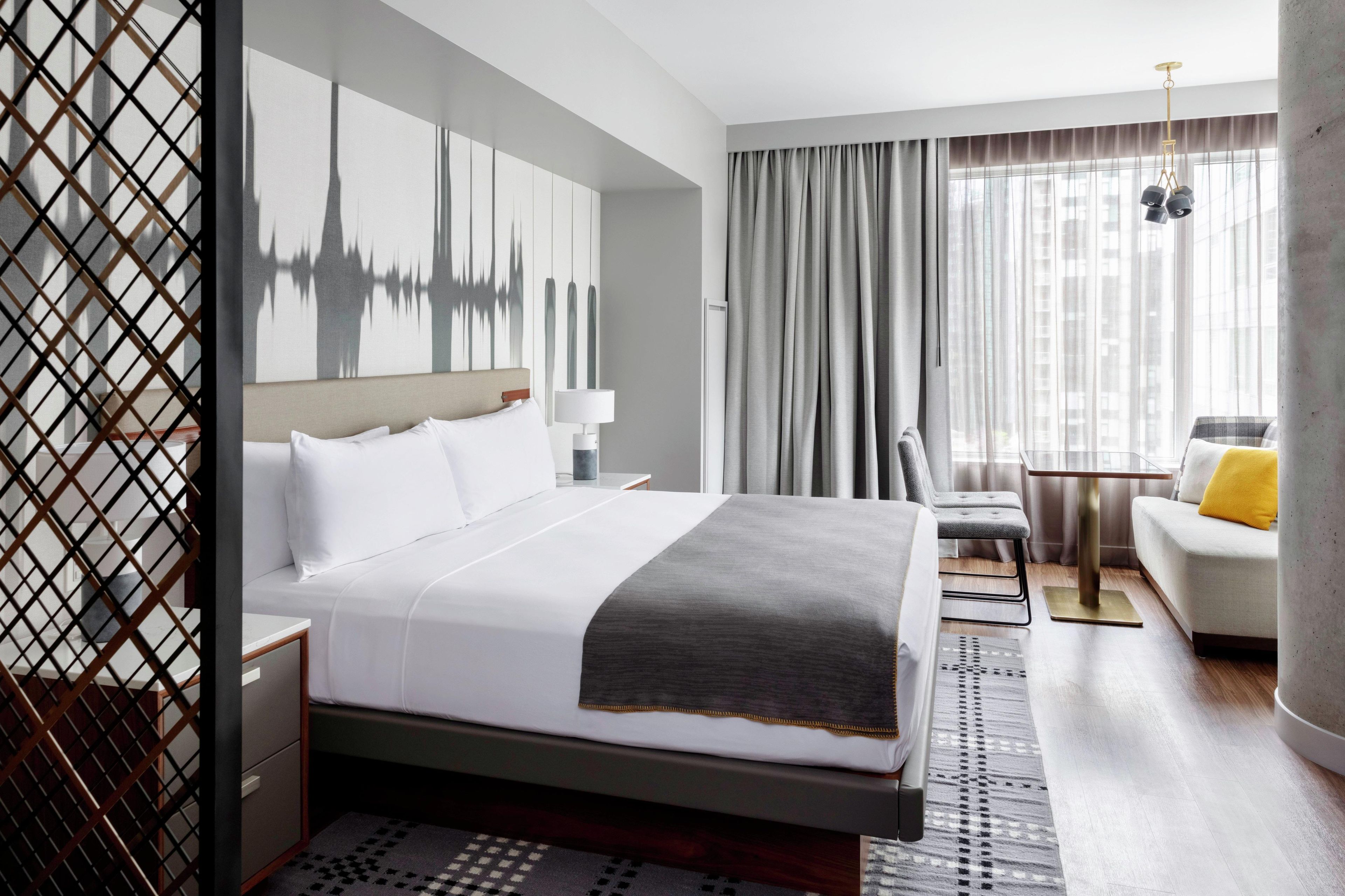 Quarto de hotel moderno com cama grande, escrivaninha, sofá e vista da cidade através de amplas janelas.