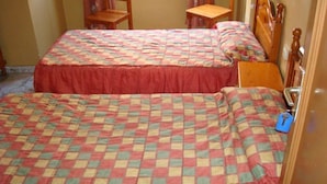 Quarto com duas camas, cada uma com colcha xadrez e saia vermelha. Há uma cadeira de madeira e uma mesinha no quarto.