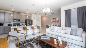 Uma cozinha moderna com armários cinza, uma ilha central com banquetas, uma área de jantar com sofá branco e mesa de centro, e uma sala de estar com uma janela grande e cortinas cinza.