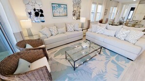 Uma sala de estar moderna com dois sofás brancos, cadeiras de vime, uma mesa de centro de vidro e um tapete estampado.