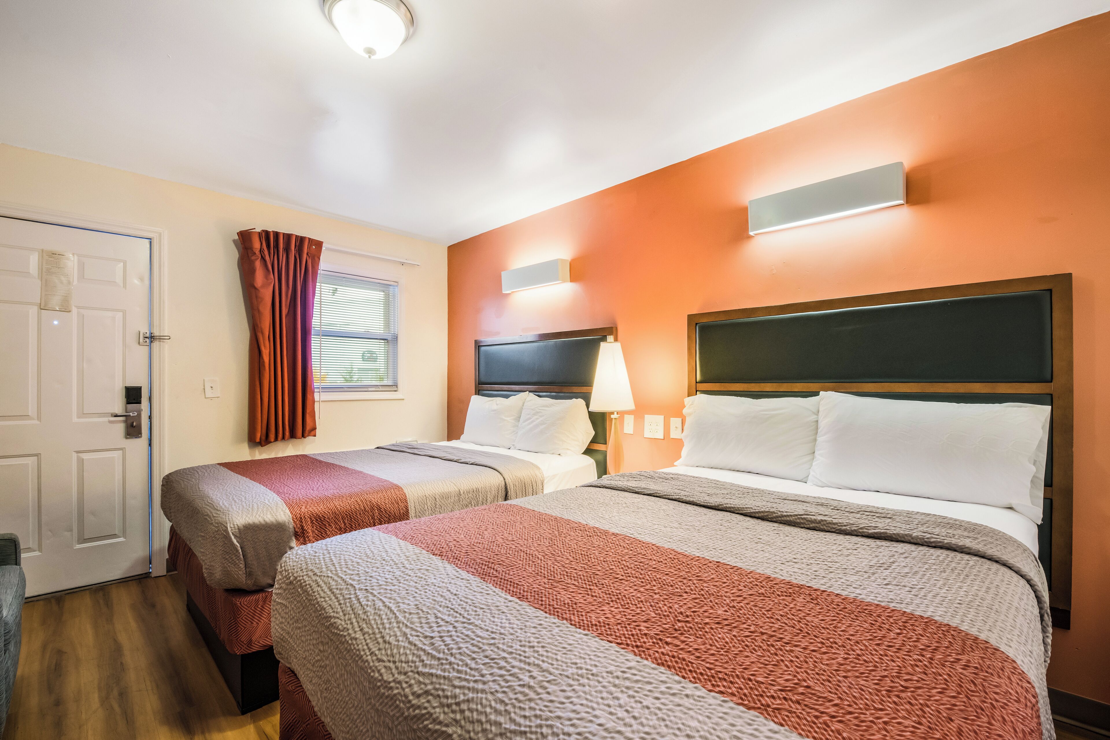 Quarto de hotel com duas camas, uma porta, uma janela com cortinas e um ponto de luz fixado na parede.