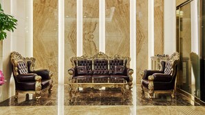 Um lobby luxuoso com parede de mármore, um sofá vermelho escuro e aconchegante, e cadeiras douradas ricamente decoradas.