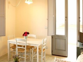 Uma sala de jantar com uma mesa e cadeiras brancas, um vaso com flores vermelhas e uma porta que dá para o exterior.