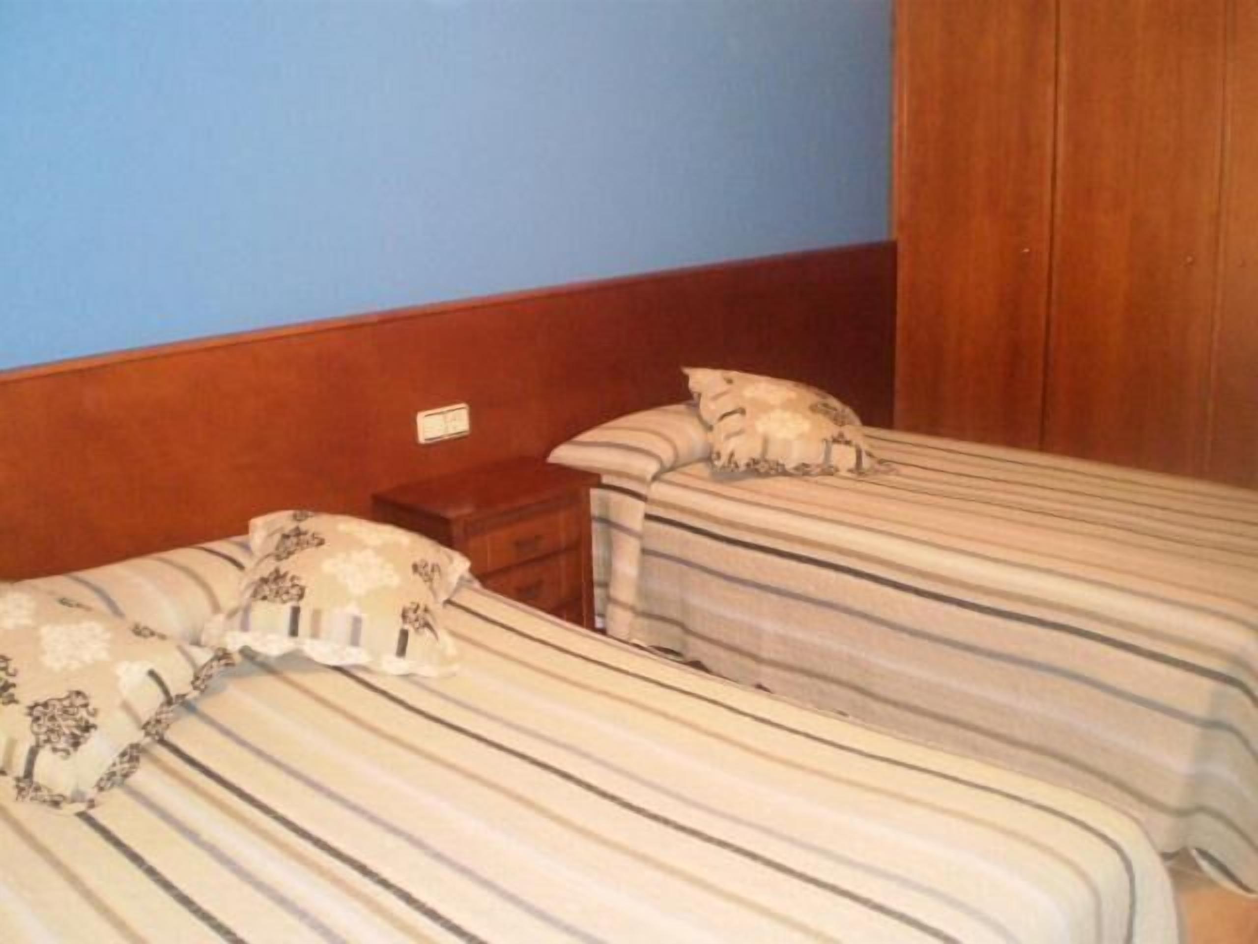 Quarto com duas camas, cada uma com roupa de cama listrada e um travesseiro. Possui cabeceira de madeira e uma pequena mesa de cabeceira com uma luminária.