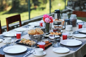 Mesa de café da manhã posta ao ar livre com diversos alimentos, um vaso de flores e uma cafeteira.