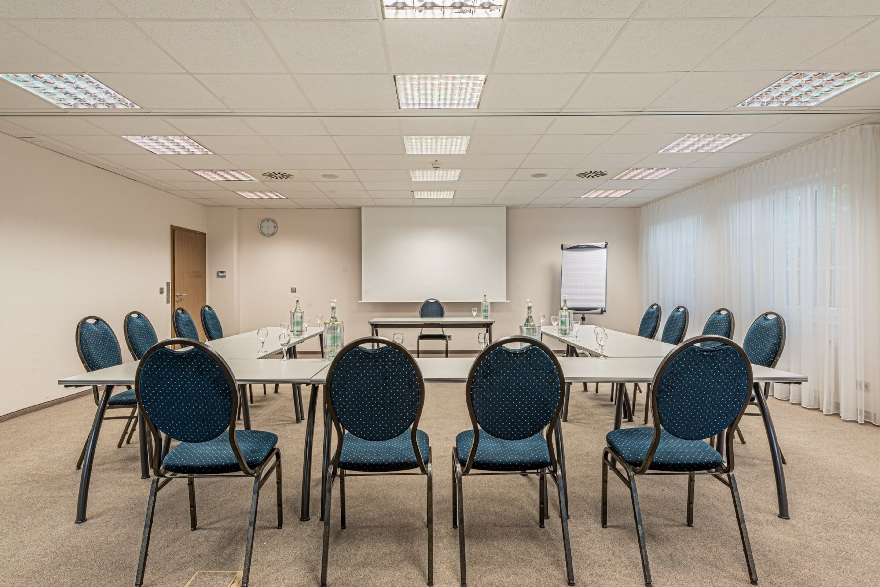 Sala de conferências com uma mesa comprida, cadeiras, tela de projetor e um relógio na parede.