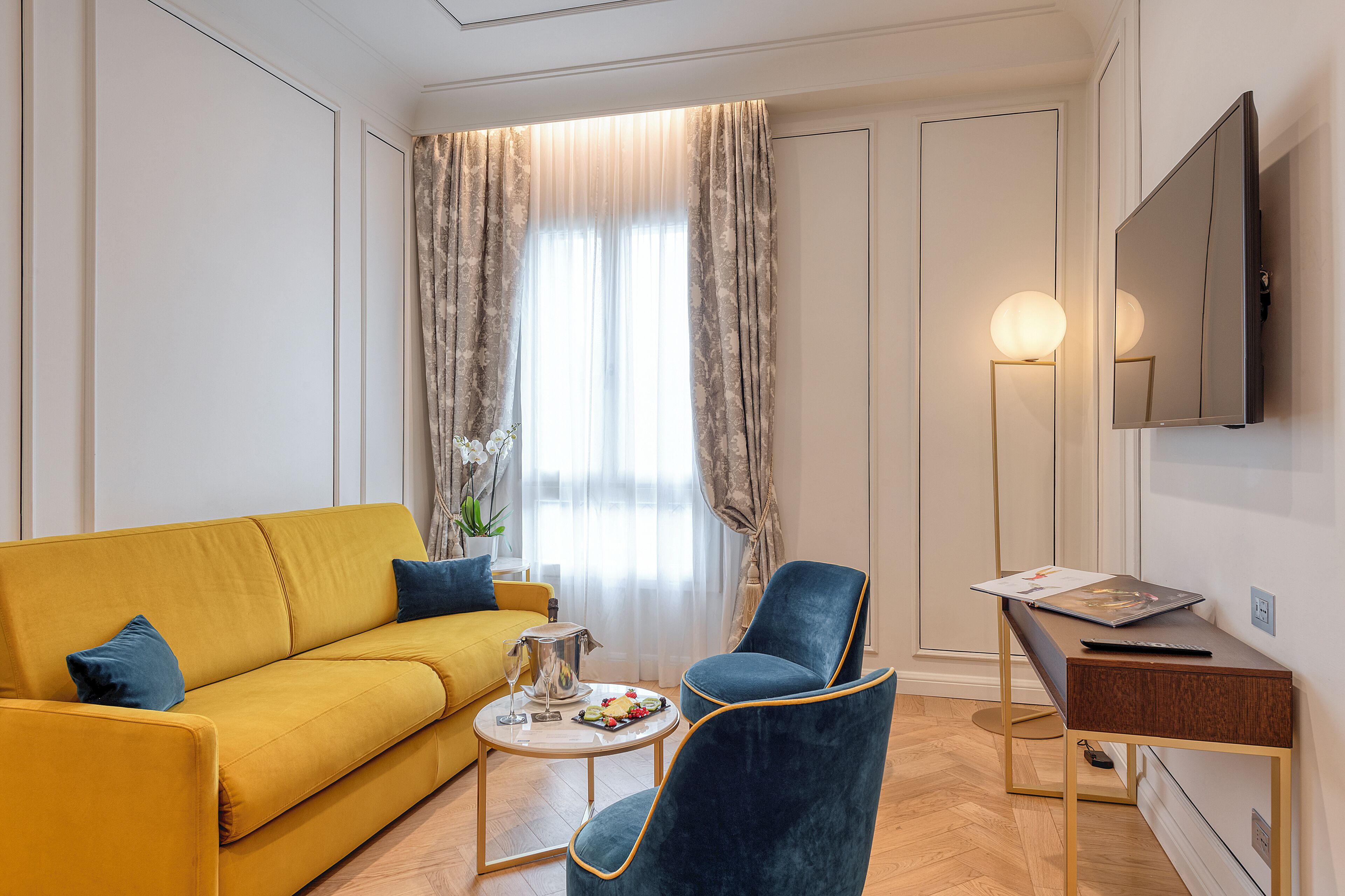 Uma sala de estar moderna com um sofá amarelo, uma poltrona azul, uma mesinha de centro e uma escrivaninha com uma televisão de tela plana.