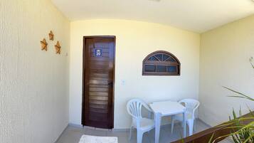 Quarto pequeno com porta de madeira identificada com o número 20, parede branca, janela pequena com persianas e duas cadeiras de plástico branco.