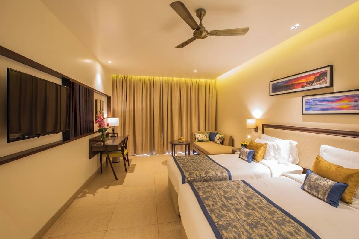 Quarto de hotel com cama, sofá, escrivaninha, cadeira, ventoinha no teto e uma janela grande com cortinas.
