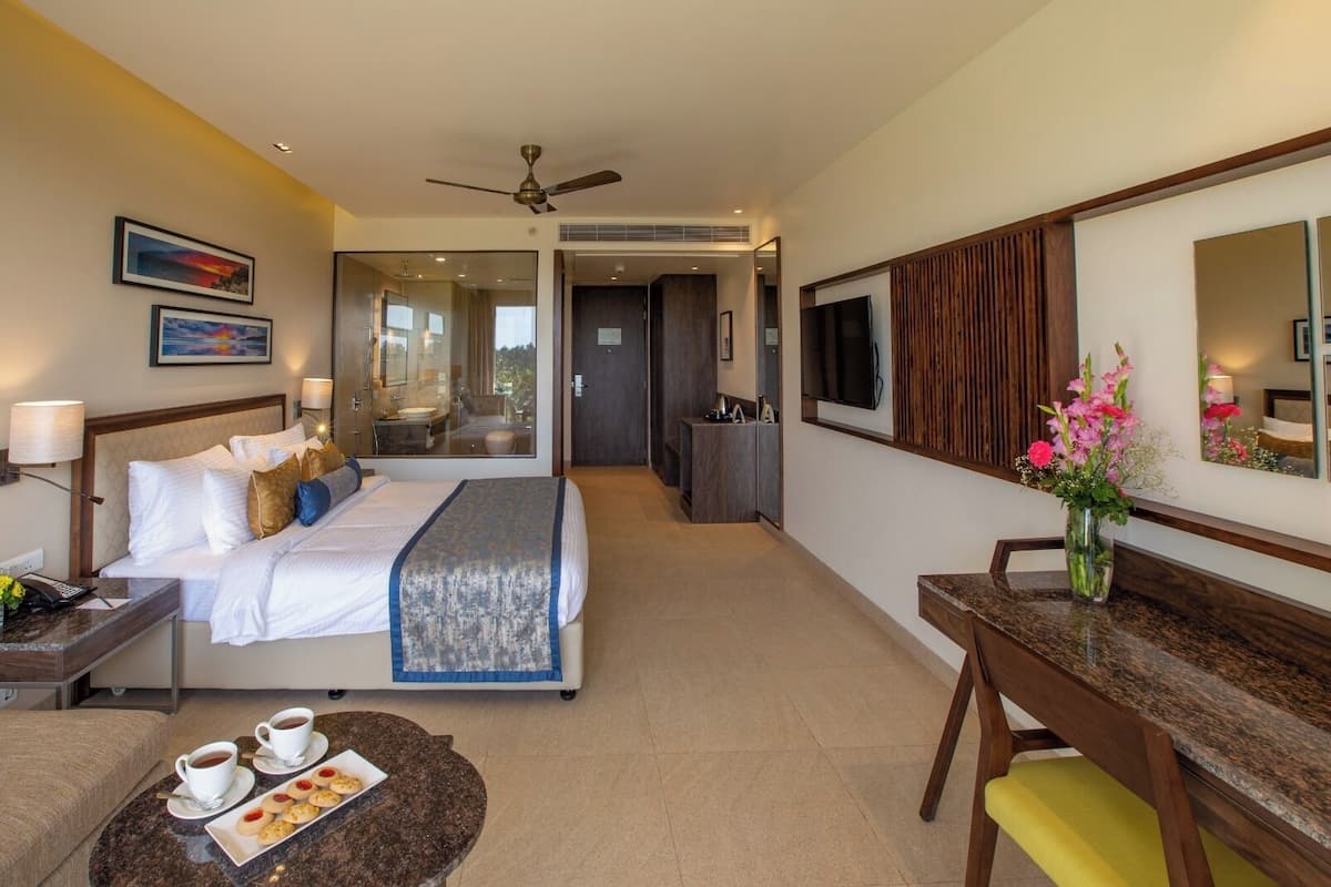 Quarto de hotel com cama, mesa de cabeceira, escrivaninha, cadeira e um espelho grande.