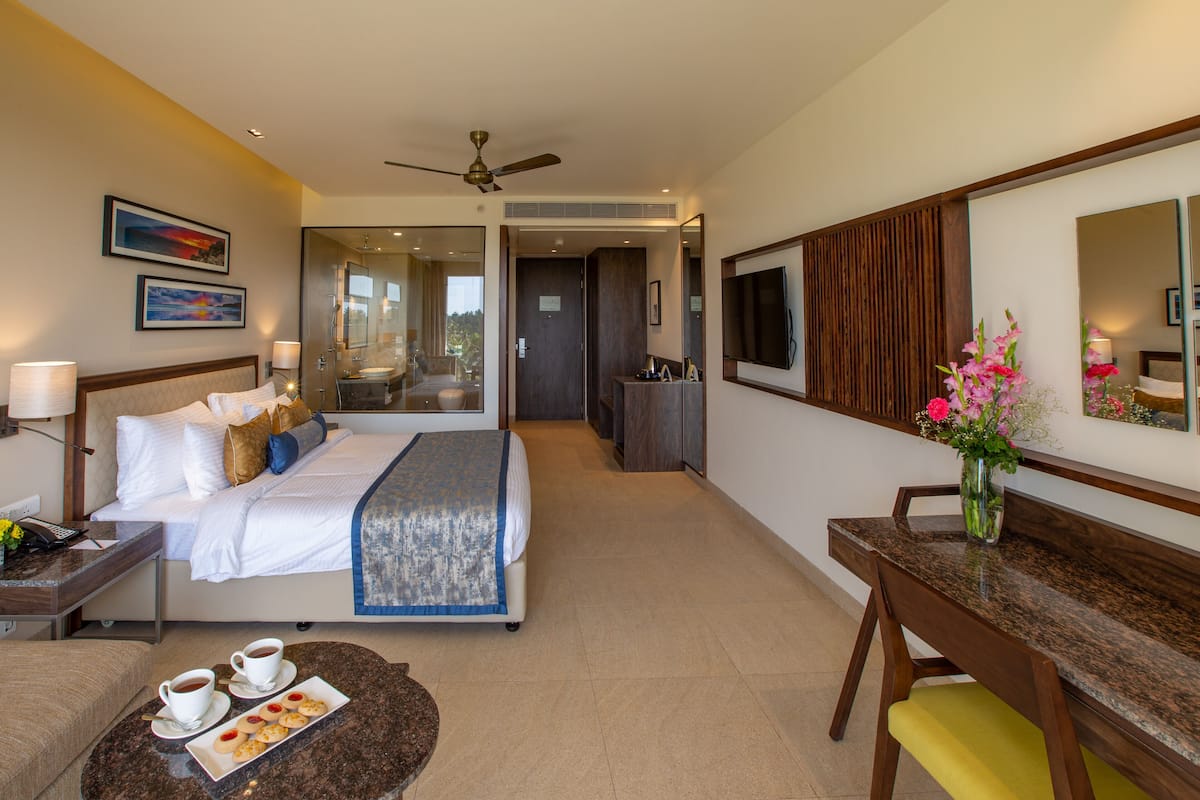 Quarto de hotel com cama, mesa de cabeceira, escrivaninha e vista para o banheiro e corredor.