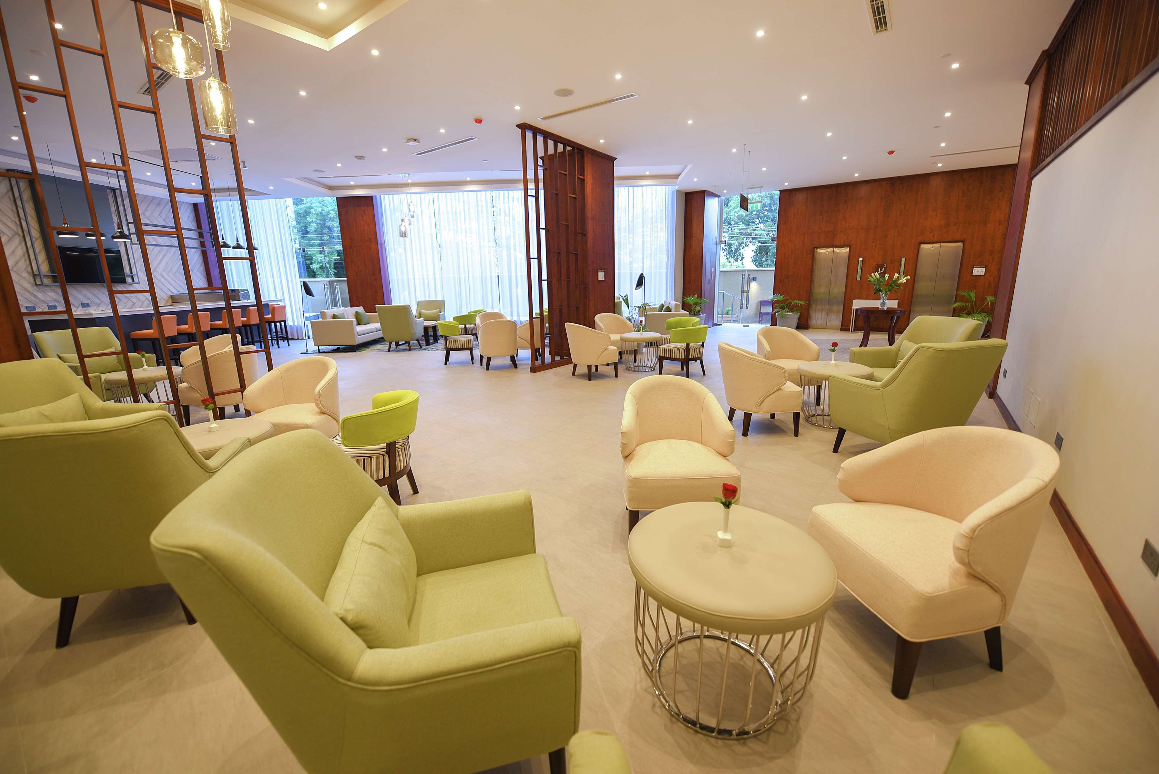 Um hall de hotel moderno com poltronas verdes e bege, uma mesa redonda central com um vaso, e uma área de cozinha visível ao fundo.