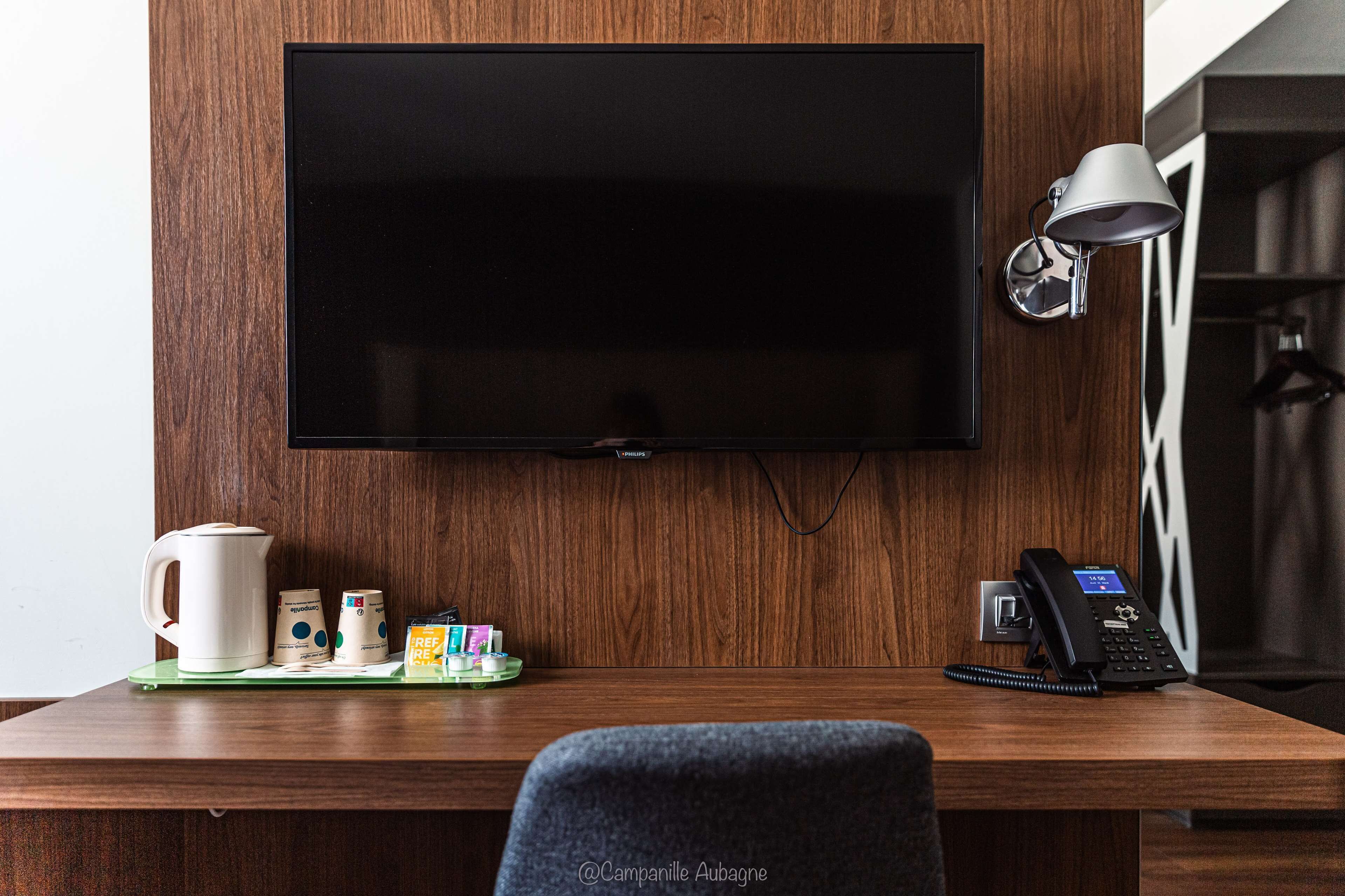 Quarto de hotel com uma escrivaninha de madeira, televisão de tela plana na parede, luminária de mesa, telefone e um descanso com cafeteira, xícaras e artigos de higiene pessoal.