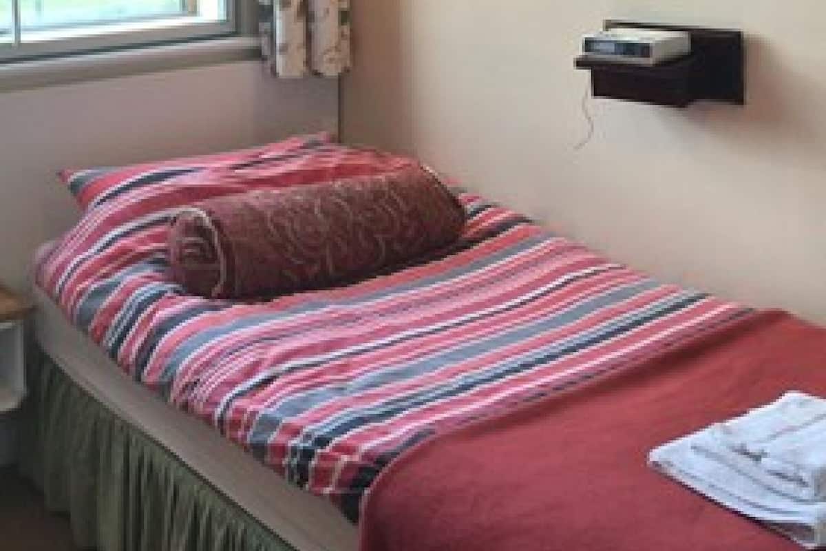 Uma cama de solteiro com colcha listrada e manta vermelha, uma janela com cortinas e um aparelho de luz fixado na parede.