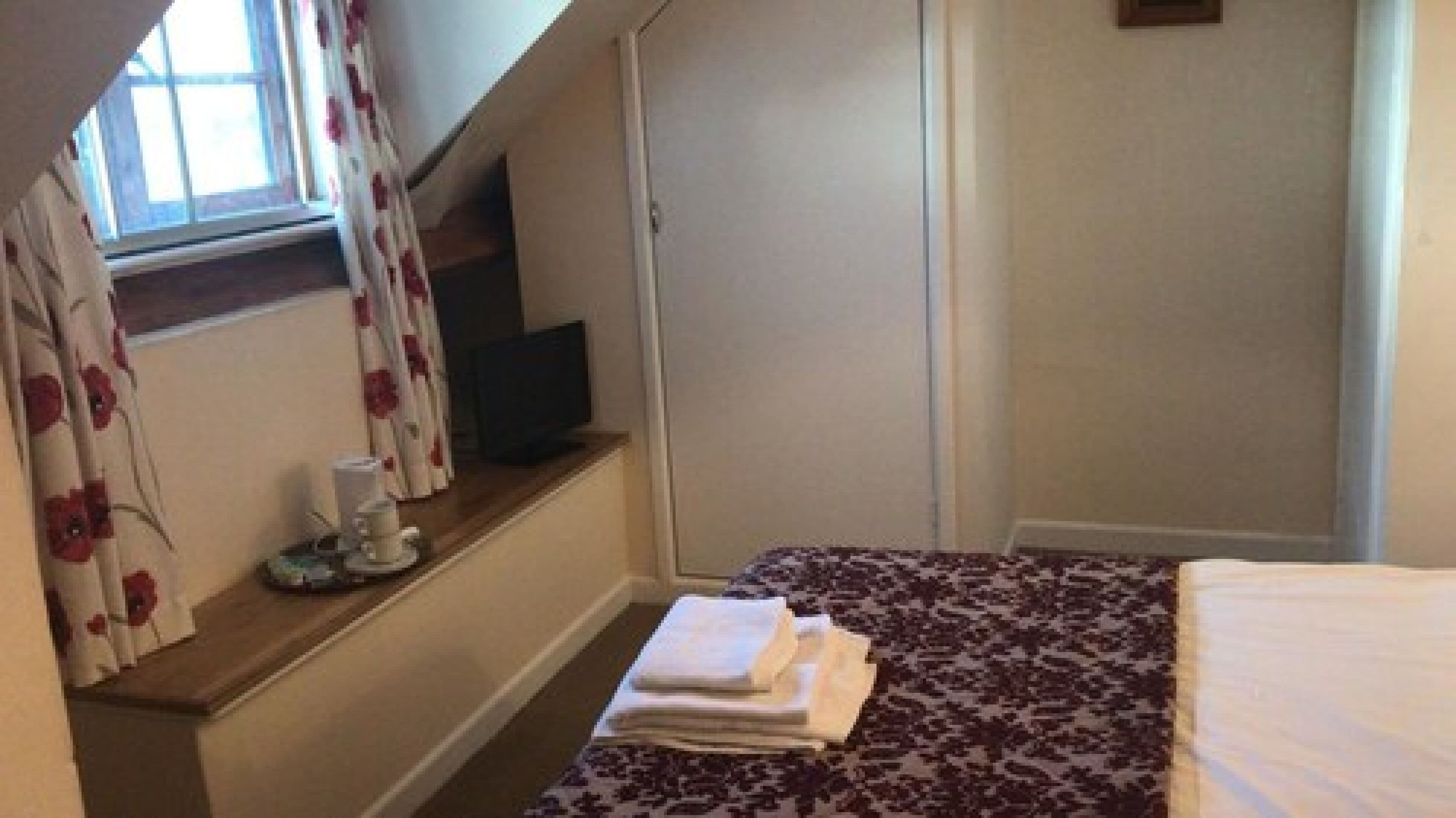 Quarto com cama, televisão em uma estante, janela com cortinas e uma mesinha com xícara e pires.