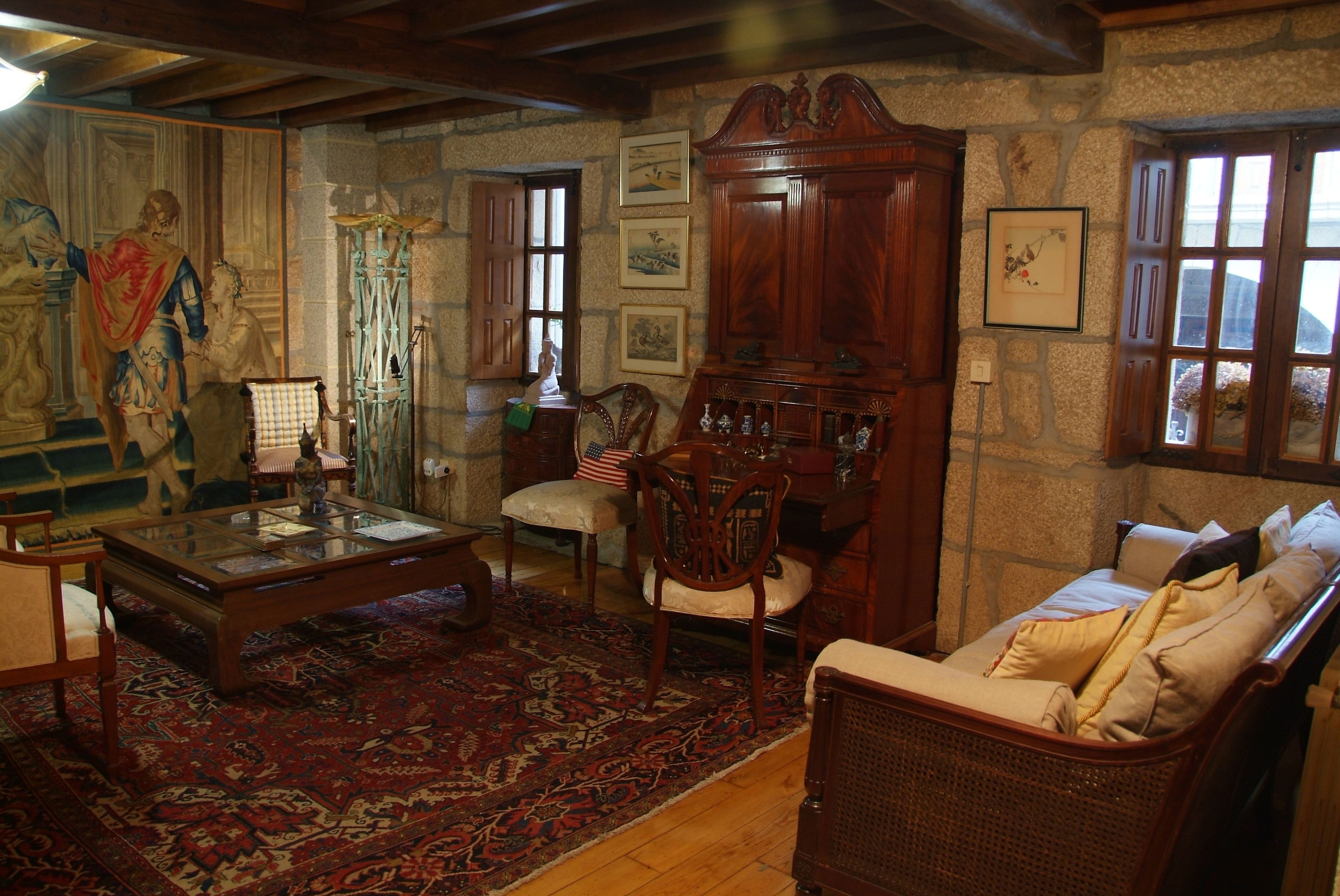 Quarto tradicional com parede de pedra, um armário de madeira, mesa de centro de vidro, sofá de vime e tapete estampado.
