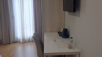 Quarto de hotel com uma escrivaninha de madeira, uma cadeira, televisão de tela plana fixada na parede, janela com cortinas finas e piso de madeira.