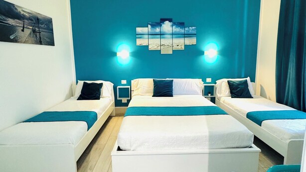 Quarto de hotel com duas camas de solteiro, uma cama de casal, uma luminária no teto, uma obra de arte na parede e uma mesa de cabeceira com abajur.