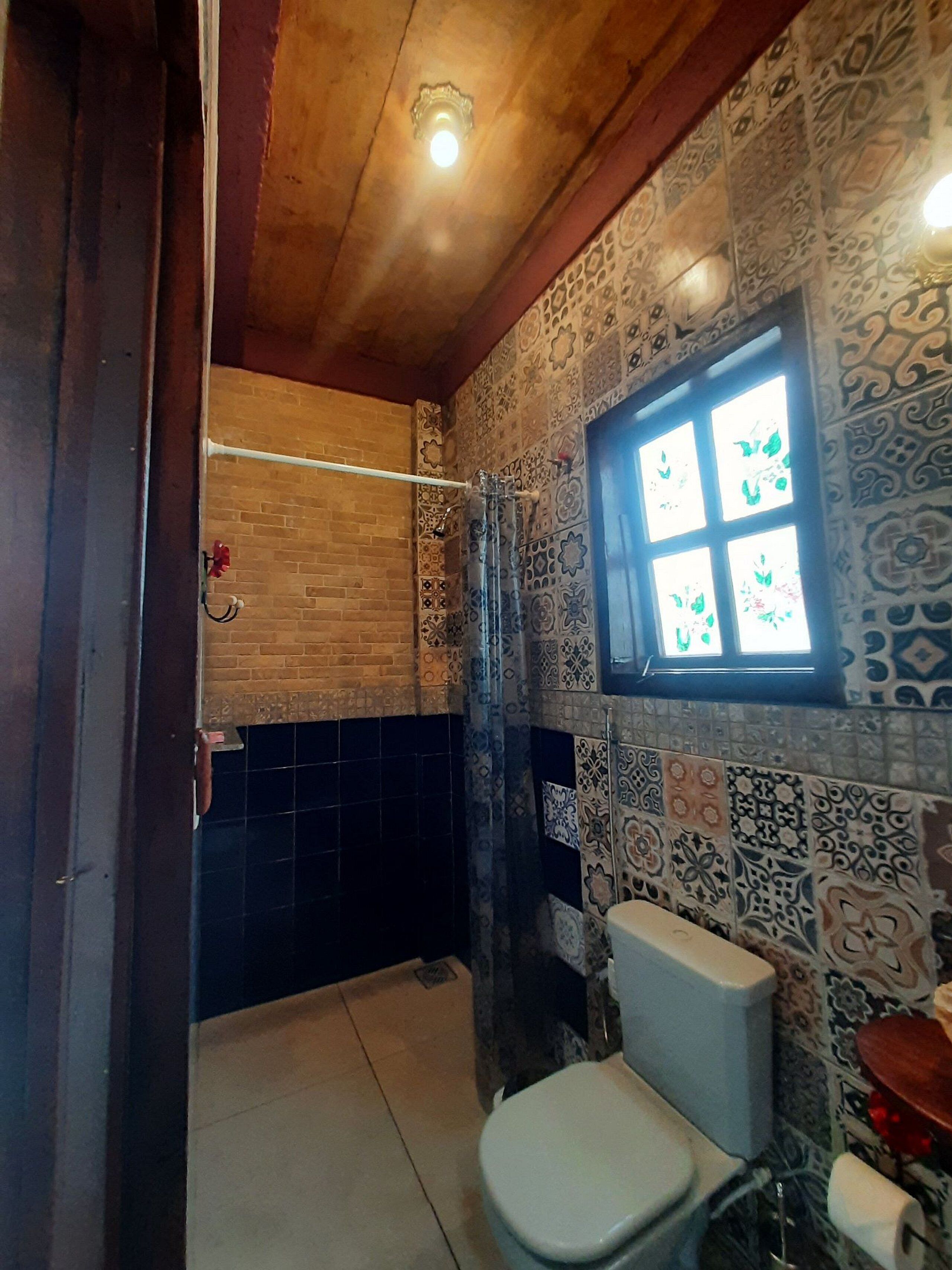 Casa de banho com chuveiro em azulejos, sanita e janela com padrão decorativo.