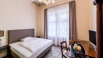 Quarto de hotel com cama, escrivaninha com cadeira, televisão e lustre.