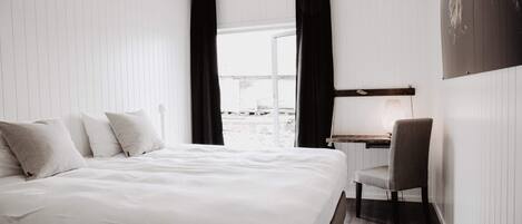 Quarto de hotel com uma cama grande, uma mesa de trabalho e uma cadeira. Há uma janela com cortinas e vista para o exterior.
