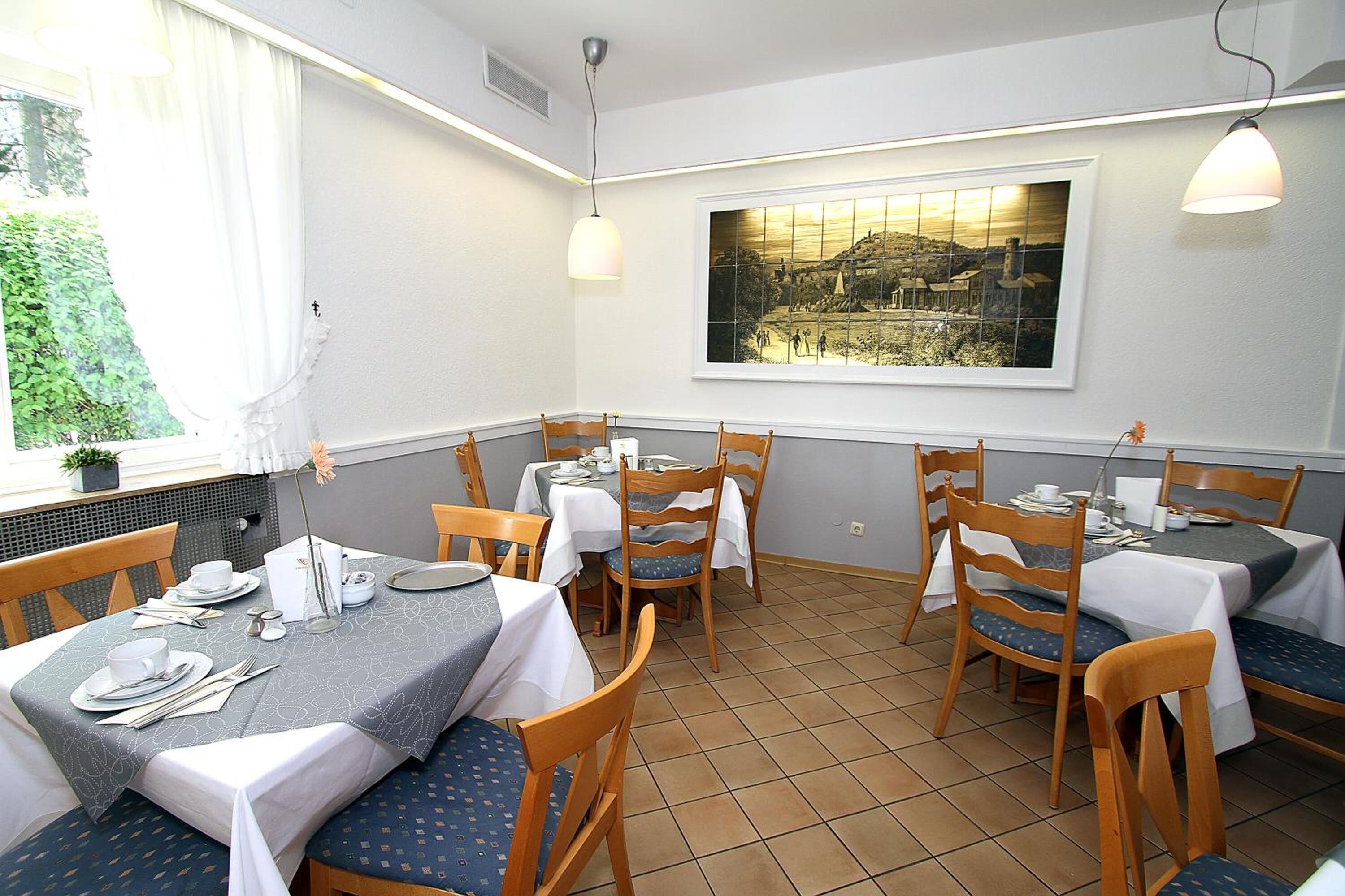 Uma sala de jantar com mesas preparadas para quatro pessoas, cadeiras de madeira com almofadas azuis, toalhas de mesa brancas e um quadro grande na parede.