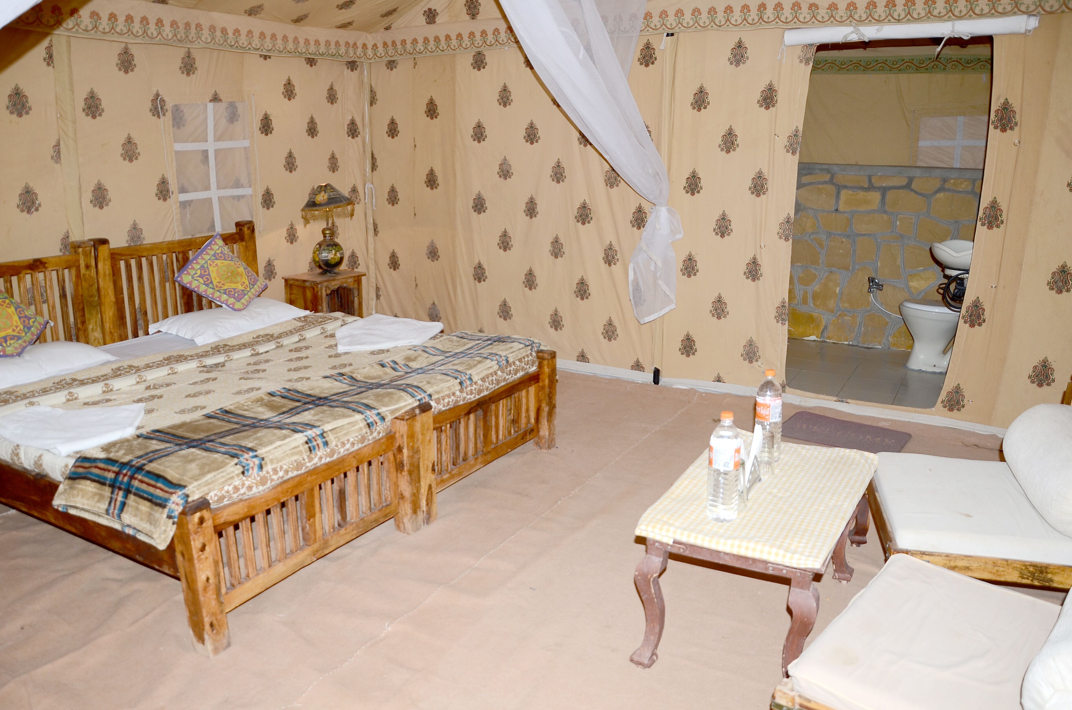 Quarto com cama de madeira, mesa de cabeceira, pequena mesa com garrafas e um banheiro visível por uma porta aberta.