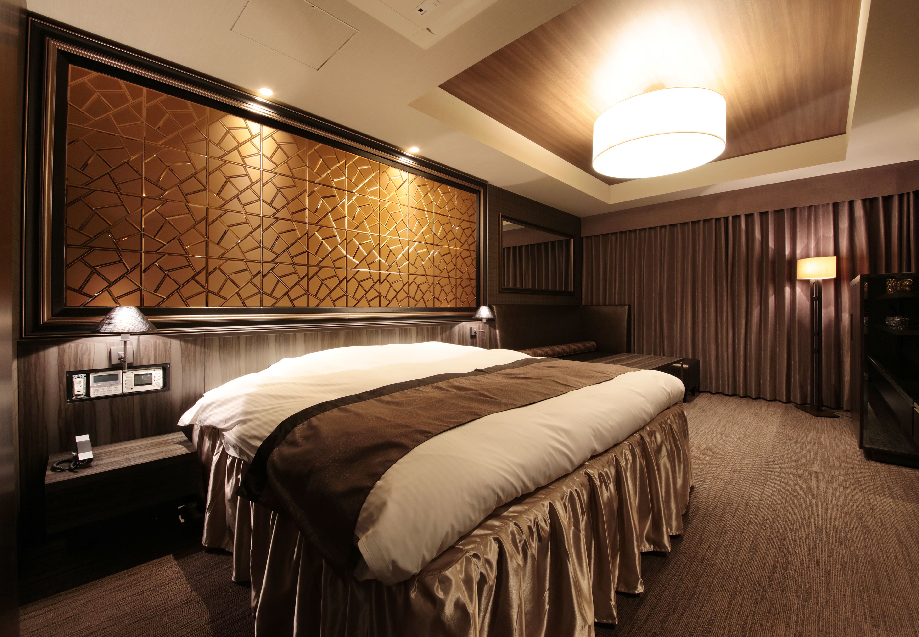 Quarto de hotel com uma cama grande, cabeceira com padrão geométrico, mesa de cabeceira com luminária e um espelho amplo.