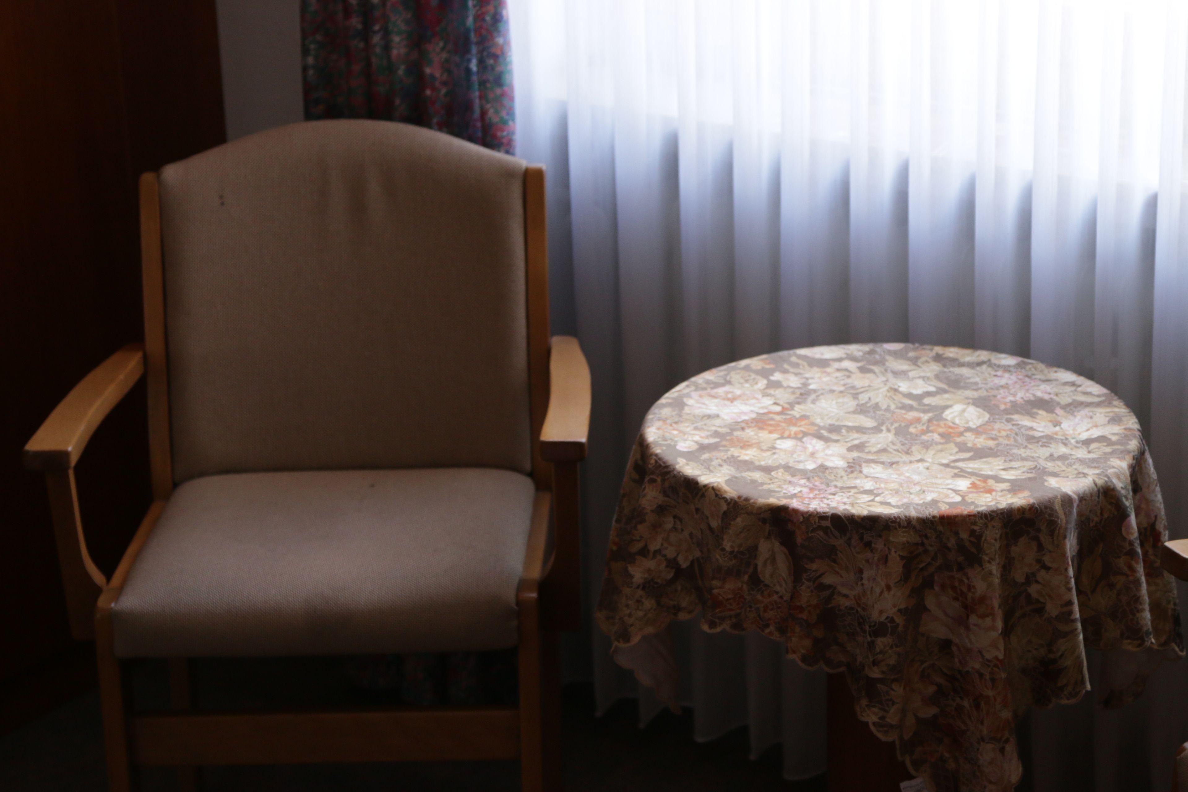 Uma cadeira de madeira e uma mesa redonda com toalha de mesa florida numa divisão com janela e cortinas.