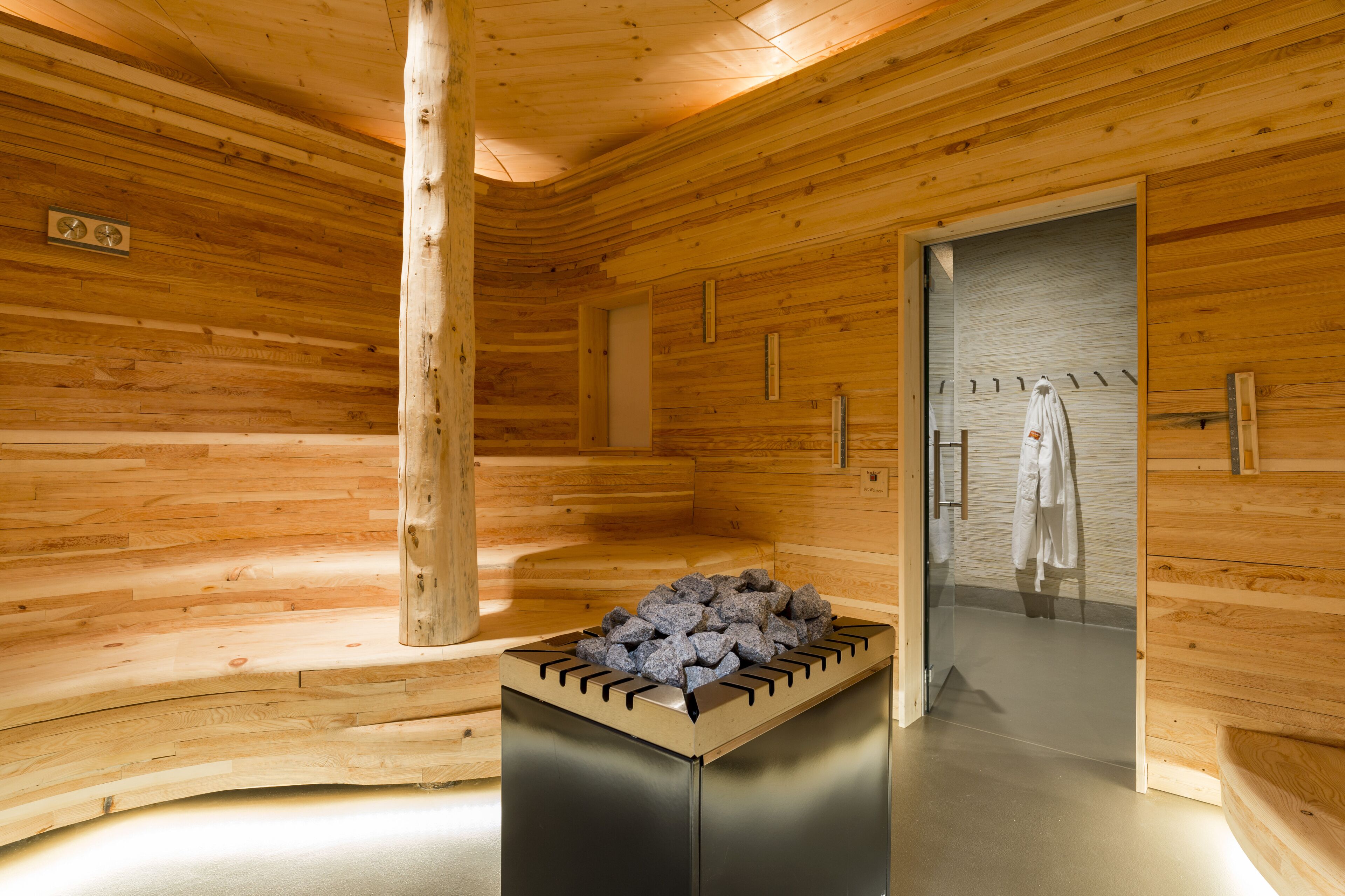 Interior de cabana de madeira com área de sauna, incluindo um banco de pedra e porta de vidro para área de chuveiro.