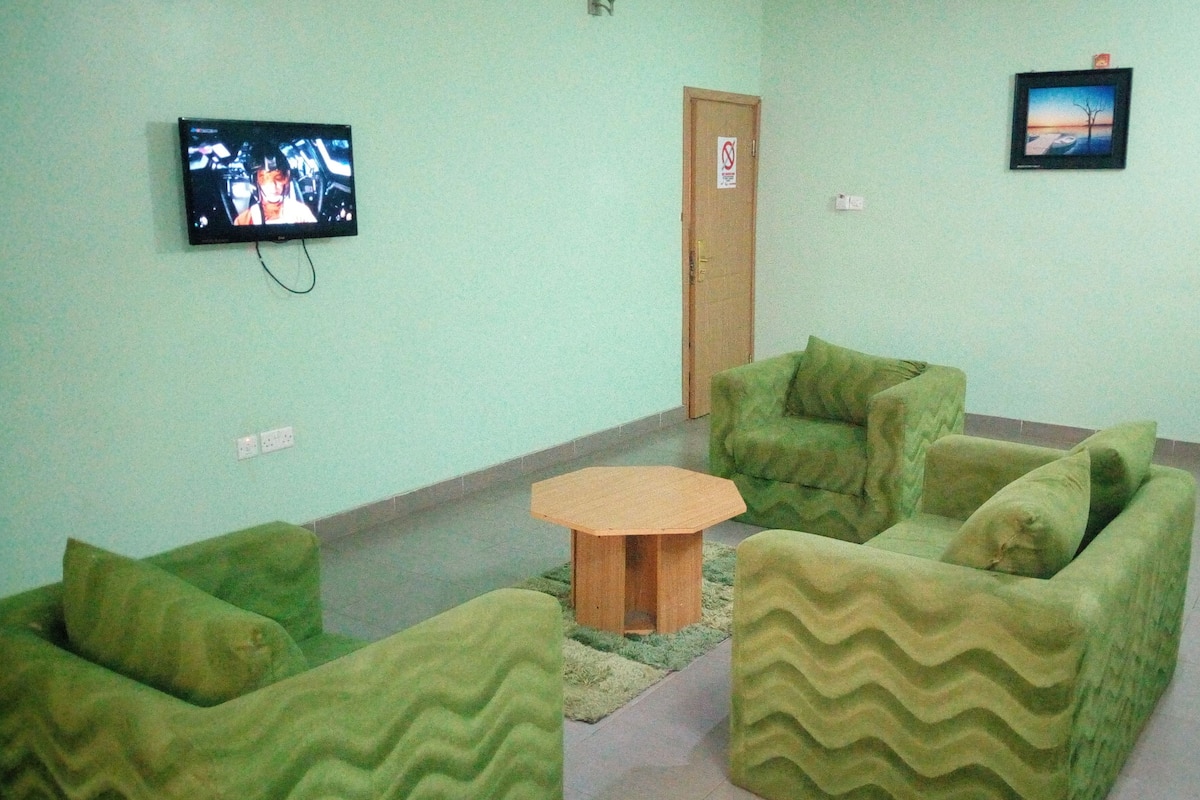 Quarto com mobiliário estofado em verde, televisão na parede e um quadro emoldurado.