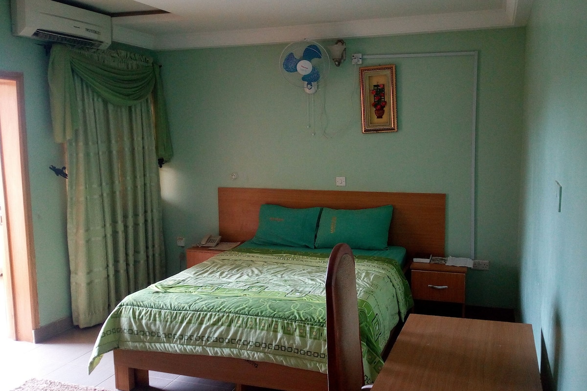 Quarto com cama, mesas de cabeceira, ar condicionado na parede, janela com cortinas e um quadro na parede.