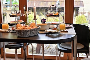 Mesa de café da manhã com diversos alimentos, incluindo pão, doces e uma cesta de frutas, com vista para o verde lá fora.