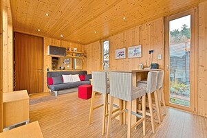 Interior acolhedor de uma cabana, com teto de madeira, um sofá, uma mesa de jantar com cadeiras e uma janela grande com vista para o exterior.