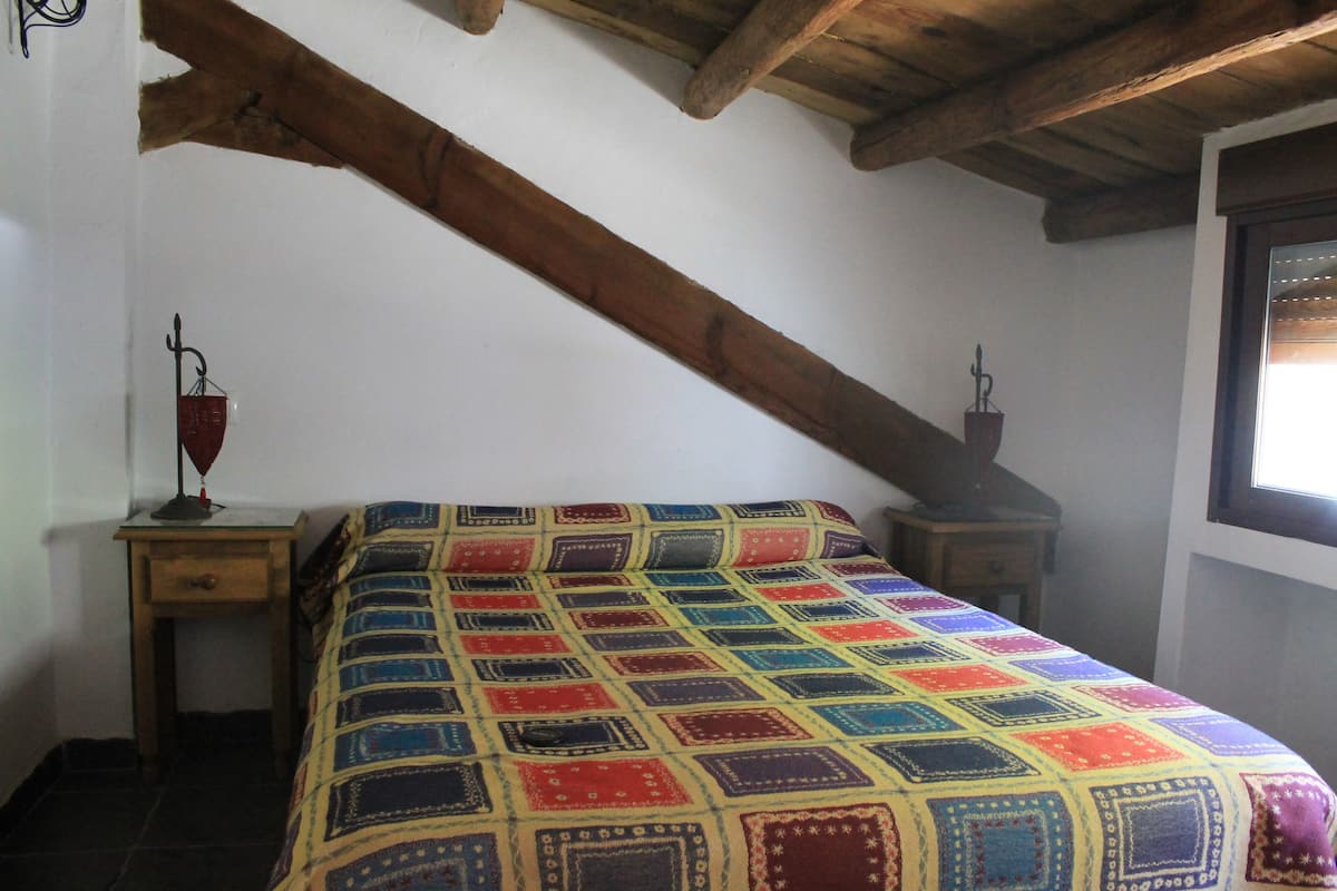 Quarto com teto de madeira, colcha colorida na cama, mesa de cabeceira com abajur e janela com persianas.