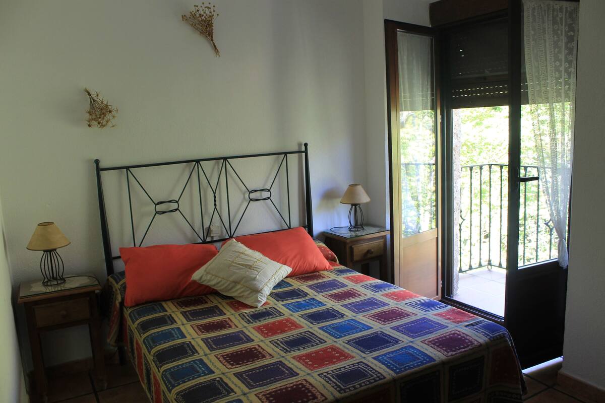 Quarto com cama, mesa de cabeceira, janela com vista para vegetação e porta que dá para uma varanda externa.