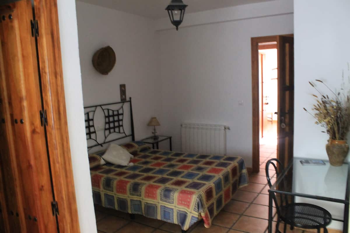 Quarto com cama, mesa de cabeceira, luminária, radiador e porta de madeira.