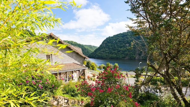 Uma casa com fachada de pedra, telhado de telhas e vista para um lago e montanhas.